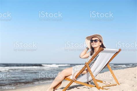 선글라스와 모자를 쓰고 바다 해변의 나무 갑판 의자에서 휴식을 취하는 행복한 갈색 머리 여자 25 29세에 대한 스톡 사진 및