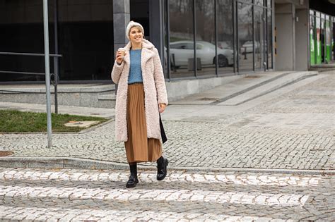 니트 모자와 인조 모피 코트를 입은 여성 휴대 전화와 컵 Uf 커피 뜨거운 음료가 횡단 보도에서 길을 건너고 도시 거리 스타일 현대 생활에서 걷는 여성 여자에 대한 스톡