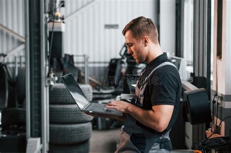 Holding Laptop In Hands Man In Uniform Is Working In The Auto Service Holding Laptop In Hands Man In Uniform Is Working In The Auto Service