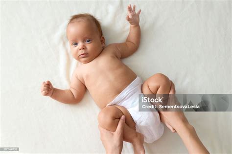 아기의 변비 가스를 배출하기 위해 갓 태어난 아이와 함께 체조를하는 엄마 아이의 다리를 움직이고 아기는 기저귀를 차고 침대에 하얀 시트에 누워 있습니다 2 5 개월에 대한