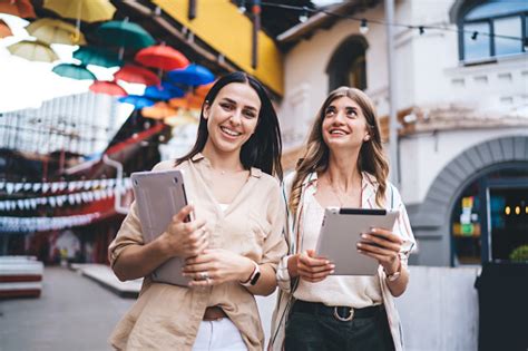 도시성에서 걷고 카메라에 미소 짓는 현대 기술을 가진 쾌활한 여성 프리랜서의 절반 길이 초상화 함께 협업을 위해 만남을 즐기는 디지털 장치를 가진 행복한 여성들 20 24