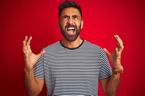 해군 줄무늬 티셔츠를 입은 젊은 잘생긴 인도 남자는 고립 된 빨간 배경에 미친 미친 소리와 공격적인 표정과 팔을 들고 고함 좌절