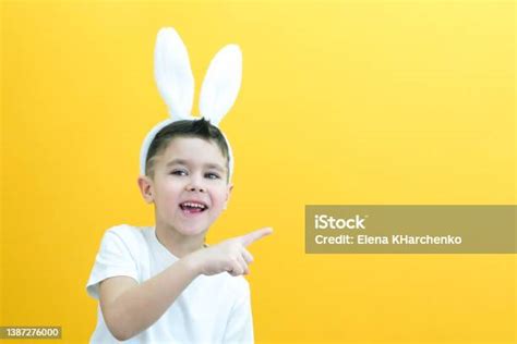 노란 배경에 그의 머리에 토끼 귀를 가진 쾌활한 소년 재미있는 행복한 아이는 빈 공간에서 손가락을 가리킵니다 텍스트 레이아웃을 위한 공간을 복사합니다 계절에 대한 스톡 사진