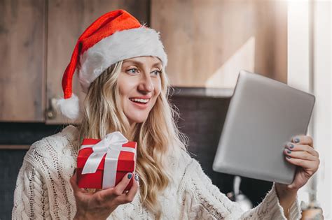 새해 화상 통화 개념 축제 크리스마스 모자를 쓴 금발 백인 소녀는 손에 태블릿을 들고 선물이 담긴 빨간 상자를 들고 미소를 짓고 화면을 바라 봅니다 새해에 대한 스톡 사진