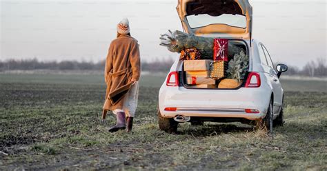 겨울 옷을 입은 여자가 해질녘 들판의 선물 상자와 크리스마스 트리로 가득 찬 차 근처를 걷는다 하늘에 복사 공간이 있는 넓은 풍경 새해 분위기의 개념 사진