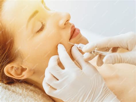 햇볕이 잘 드는 의료 센터에서 눈을 감고 미용 주사를 받는 아름다운 여성 미용사 의사가 주사기로 여성 얼굴에 미용 시술을 하고 있습니다 미용 의학 및 수술 개념
