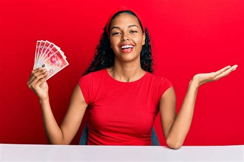 행복한 미소와 승자 표정으로 성취를 축하하는 테이블에 앉아 있는 이스라엘 셰켈을 들고 있는 젊은 아프리카계 미국인 소녀 18 19세에 대한 스톡 사진 및 기타 이미지