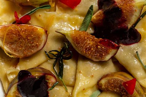 오리 필레 만두에 무화과 체리 소스 식용 보라색 꽃잎 로즈마리 체리 소스를 얹었습니다 음식은 타원형의 흰색 세라믹 접시에 놓여 있습니다 접시는 어두운 패브릭 배경 커피