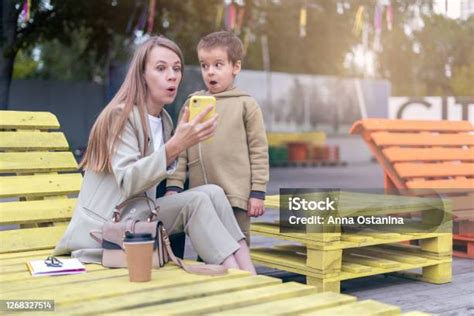 유치원 생 아들과 재미와 놀란 젊은 엄마는 스마트 폰을 사용하여 벤치에 앉아 함께 재미를 어머니와 어린 소년 아이 즐길 주말