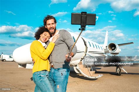 공항 피스트에 주차된 전용 비행기 앞에서 셀카 봉에 장착된 스마트폰으로 셀카를 찍고 있는 풍부한 다민족 커플 Monopod에