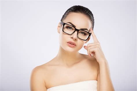 검은 머리가 뒤에 고정 된 화려한 소녀 큰 눈 두꺼운 눈썹 및 안경을 쓰고 회색 스튜디오에서 포즈를 취하는 벌거 벗은 어깨