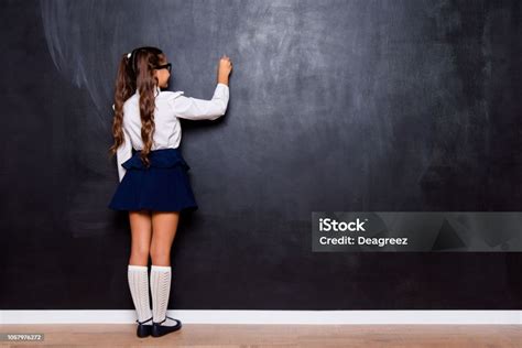 후면 보기 다시 좋은 천재의 흰 정장 블라우스 셔츠 곱슬 묶은 머리와 함께 사랑 스러운 작은 소녀는 칠판에 쓰는 블루 치마 짧은 검은 배경 위에 절연 교육에 대한 스톡 사진