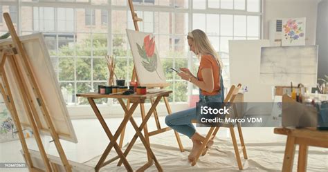 그림이있는 스튜디오의 아티스트는 소셜 미디어 게시물에 대한 전화 또는 잠재적 인 구매자의 텍스트를 확인하십시오 여성은 캔버스에서 예술로 휴식을 취하고 스마트 폰을 사용하여