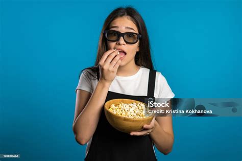 공포 영화를보고 파란색 스튜디오 배경에 팝콘을 먹는 3d 안경에 젊은 아시아 여성 때문에 영화에서 예기치 않은 음모의 무서운 소녀 갈색 머리에 대한 스톡 사진 및 기타