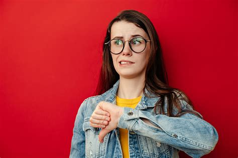 안경을 쓴 불만 스러운 여자는 비승인 기호를 보여줍니다 엄지 손가락을 유지 싫어 표현 불만에 얼굴을 찡그린 얼굴 데님 재킷을 입고 빨간 배경 위에 고립 바디 랭귀지