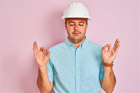 고립 된 분홍색 배경 위에 서있는 보안 헬멧을 착용한 젊은 건축가 남자는 손가락으로 명상 제스처를하고 눈을 감고 미소지으며 휴식을 취합니다 요가 개념입니다 개념에 대한 스톡