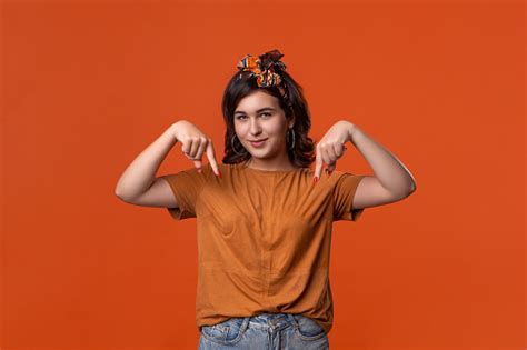 예쁜 갈색 머리 여자 에 티셔츠와 아름 다운 머리띠 가리키는 그녀의 손가락 아래로 오렌지 배경 위에 고립 된 광고를 위한 장소입니다 T 셔츠에 대한 스톡 사진 및 기타