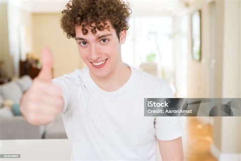 젊은 잘생긴 남자를 입고 흰색 T 셔츠를 하 고 행복 한 엄지손가락을 손으로 제스처 카메라를 보고 성공을 나타내는 식을 승인 합니다 T 셔츠에 대한 스톡 사진 및 기타