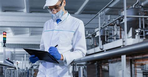 모자 마스크 및 흰색 코트의 기술자가 생산 공장에 서서 시트에 펜으로 메모를합니다 검사관은 낙농 공장에서 표시기를 제거합니다 엔지니어는 생산에 대한 통계를 유지합니다