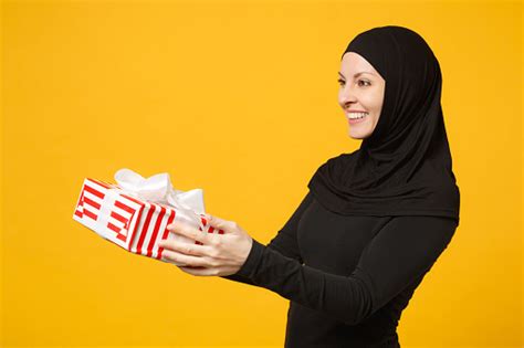 웃는 젊은 아라비아 무슬림 여자 히 잡 블랙 옷 노란 벽 배경 스튜디오 초상화에 고립 된 선물을 가진 손에 선물 상자에 개최