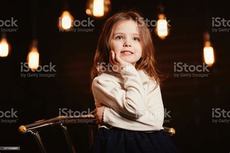 작고 사랑스러운 웃는 소녀는 손으로 얼굴을 만지고 어두운 배경으로 카메라를 보고 있다 레이스 블루 드레스와 니트 스웨터를 입은 아이가 금속 의자에 앉아 있다 행복한 어린
