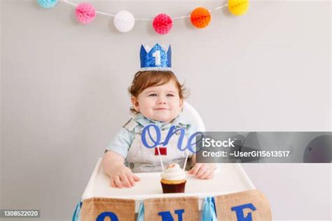 집에서 첫 번째 생일을 축하하는 파란색 왕관에 귀여운 미소 백인 아기 소년 토퍼 단어 One맛있는 컵 케이크 디저트를 먹는 높은 의자에 앉아 있는 어린이 유아 생일 컨셉
