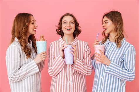 핑크 배경에 고립 된 행복한 수면 중 종이 컵에서 미소와 차가운 탄산음료를 마시는 다채로운 줄무늬 잠옷에 좋은 분위기를 가진 세 가지 콘텐츠 여성 3 명에 대한 스톡 사진