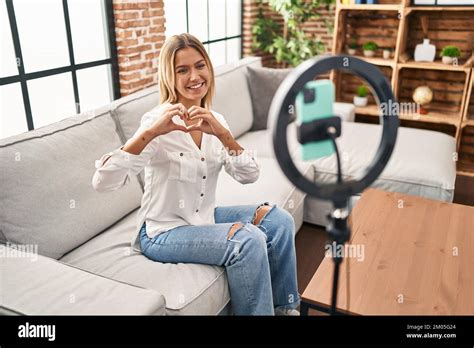 Young Blonde Woman Recording Vlog Tutorial With Smartphone At Home Smiling In Love Doing Heart