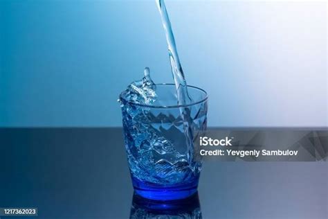 깨끗한 물은 검은 탁자 위에 서 있는 유리에 부어넣습니다 파란색 배경에 검은 테이블에 흐르는 물이있는 파란색 유리 0명에 대한