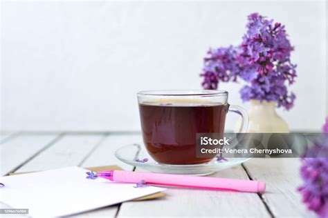 라일락 꽃 투명한 차 한 잔 접시 흰 화병 화이트 및 크래프트 페이퍼 흰색 나무 테이블에 분홍색 펜이 있는 섬세한 아침 차 테이블 세팅 Mockup 공간을 복사합니다 봄