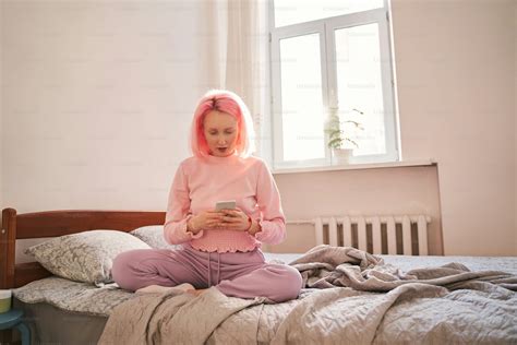 가제트와 함께 즐기기 분홍색 머리를 가진 여자가 침대에 앉아 아침에 스마트폰에 무언가를 타이핑하는 모습 스톡 사진 사진