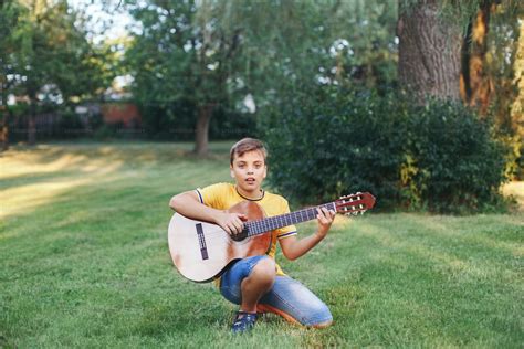 야외에서 기타를 연주하는 십대 초반의 소년을 듣기 어렵습니다 귀에 보청기를 착용한 아이는 공원에서 음악을 연주하고 노래를