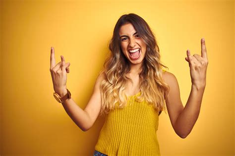 노란색 고립 된 배경 위에 티셔츠를 입고 젊은 아름다운 여성은 손을 위로 바위 기호를하고 미친 표정으로 외칩니다 음악 스타