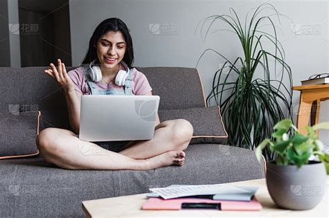 一名年轻的拉丁女学生，戴着耳机，坐在家里的沙发上用笔记本电脑进行视频通话或在线课程