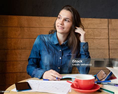 카페에 앉아 있는 파란색 데님 셔츠를 입은 쾌활한 소녀가 펜이 달린 메모장에 메모를 합니다 종이의 테이블 시트에 케이스안경 접착제 테이프 펠트 팁 펜 책 빨간 머그잔 18