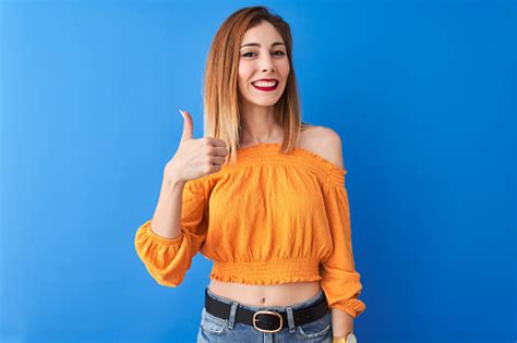 아름 다운 빨간 머리 여자 오렌지 캐주얼 티셔츠 를 입고 고립 된 파란색 배경 위에 서 행복 한 엄지 손가락 을 하 고 손으로 제스처 성공을 보여주는 카메라를 보고 표정을