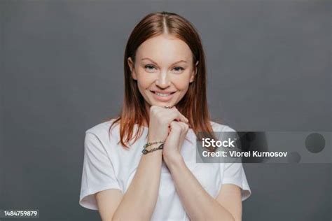 수평 주사 기쁘게 아름 다운 여자의 캐주얼 흰색 T 셔츠를 착용 좋은 분위기 어두운 회색 스튜디오 배경 포즈에 부드럽게 손을 아래 턱 미소를 유지 합니다 긍정적인 감정 30