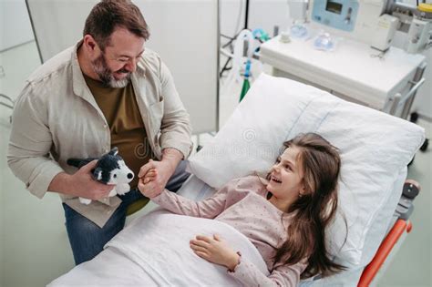 Father Sitting Beside Daughter Lying On Hospital Bed Playing With