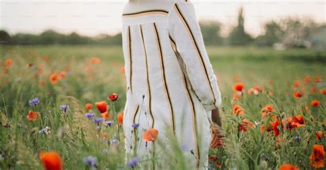 모자를 쓴 세련된 소녀는 여름 초원의 일몰 빛 속에서 야생화를 걷고 있다 시골에서 양귀비와 수레국화 사이를 걷고 있는 린넨 드레스를 입은 젊은 여성 시골의 느린 삶