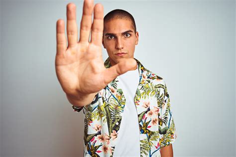 휴일에 젊은 잘 생긴 남자는 흰색 배경위에 하와이 셔츠를 입고 손바닥으로 노래를 멈추고 있습니다 얼굴에 부정적이고 심각한 제스처로 경고 표현 거절에 대한 스톡 사진 및 기타