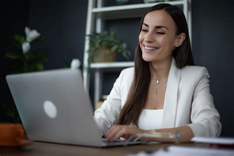 흰색 블레이저를 입은 전문직 여성이 현대적인 사무실 공간의 책상에서 노트북으로 작업하면서 미소를 짓고 있습니다 이 이미지는 생산성 자신감 긍정적인 일과 삶의 균형을