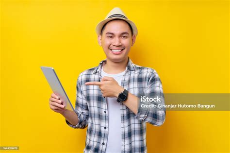 캐주얼 옷 모자를 쓴 웃는 여행자 관광객 아시아 남자가 노란색 배경에 고립된 태블릿 Pc 컴퓨터를 손가락으로 가리키고 있다 주말에 여행하는 승객 항공 비행 여행 개념 30