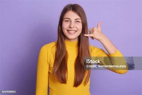 카메라를 직접 바라보고 라일락 스튜디오 벽에 서서 손가락으로 전화 제스처를 보여주는 웃는 여성의 사진 전화하세요 여성 드레스 캐주얼 옐로우 셔츠 긴 아름다운 머리를 갖는