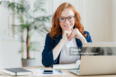 기쁘게 쾌활 한 빨간 헤어 D 여성 경제학자는 사무실 인테리어에 포즈 공식적인 옷을 입고 비즈니스 영역에서 작동 행복 한 표현이 회사를 소유 하 고 금융 시작 프로젝트를