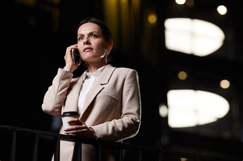 커피 컵을 들고 외국 비즈니스 파트너와 야간 통신을 위해 휴대 전화를 사용하는 베이지 색 재킷에 심각한 자신감 중년 여성 경영자에 대한 스톡 사진 및 기타 이미지 Istock