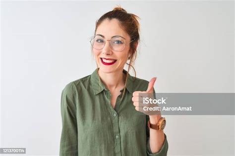 녹색 셔츠와 안경을 입고 고립 된 흰색 배경 위에 서있는 빨간 머리 여성은 행복한 엄지 손가락으로 손을 들고 제스처를 취합니다