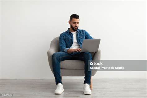 온라인 작업 연구 또는 의사 소통을 위해 노트북을 사용하는 아랍 남자의 전체 길이 안락 의자 흰색 스튜디오 벽에 앉아 앉음에