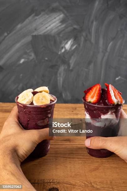 딸기와 바나나를 곁들인 플라스틱 컵에 브라질 냉동 아사이 베리 아이스크림 스무디를 들고 있는 남자 나무 책상과 과일이 있는 회색 여름 배경에 있습니다 메뉴 및 소셜 미디어에