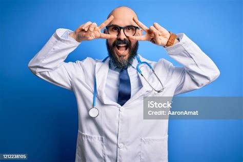 잘 생긴 대머리 의사 남자 수염 을 입고 안경과 청진기를 통해 푸른 배경 얼굴에 손가락으로 평화 기호를하고 승리를 보여주는
