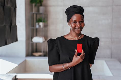 검은 터번과 검은 드레스를 입은 수줍은 아프리카 젊은 여성이 신용 카드를 들고 집에 서서 직업 경력 임금에 만족하며 카메라를 행복하게 바라보고 있습니다 성공한 사람들 재정적
